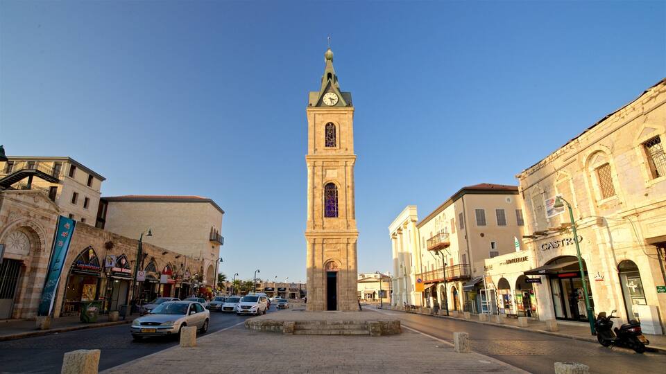 Uma praça histórica com uma torre do relógio imponente, rodeada por edifícios tradicionais e um céu azul límpido.