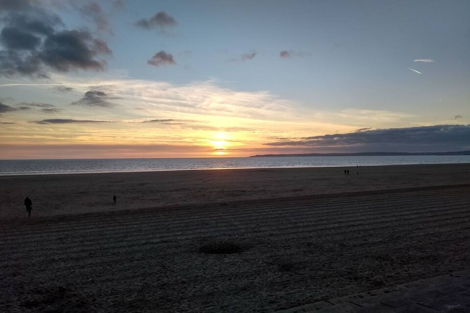 Praia ao pôr do sol, com o sol próximo ao horizonte, algumas pessoas caminhando e céu claro com nuvens.