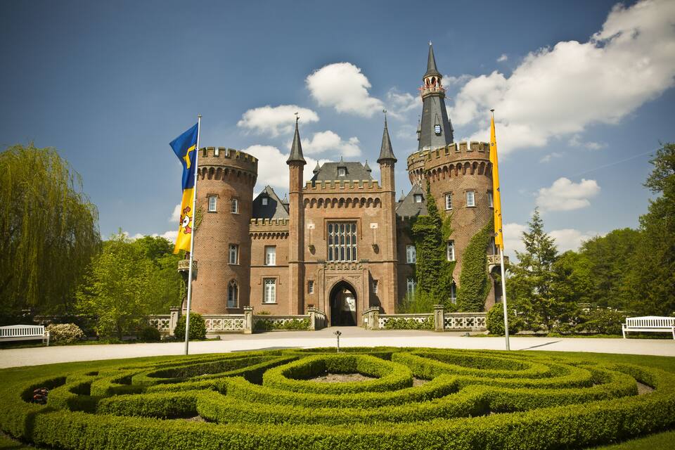 Um castelo com várias torres e bandeiras, rodeado por um jardim bem cuidado e árvores.