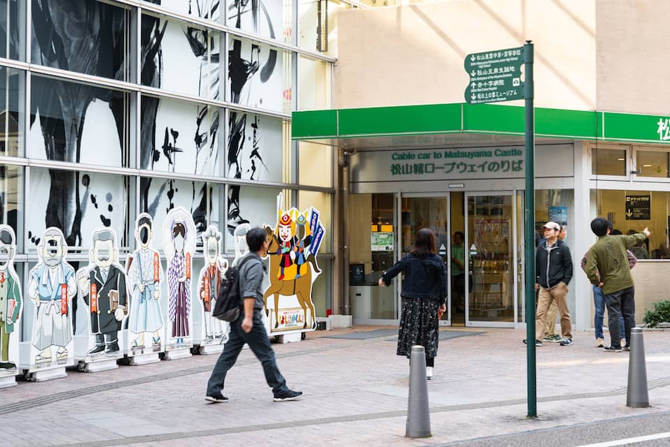 Um edifício com toldo verde e uma placa em japonês. Há figuras recortadas de pessoas na parede.