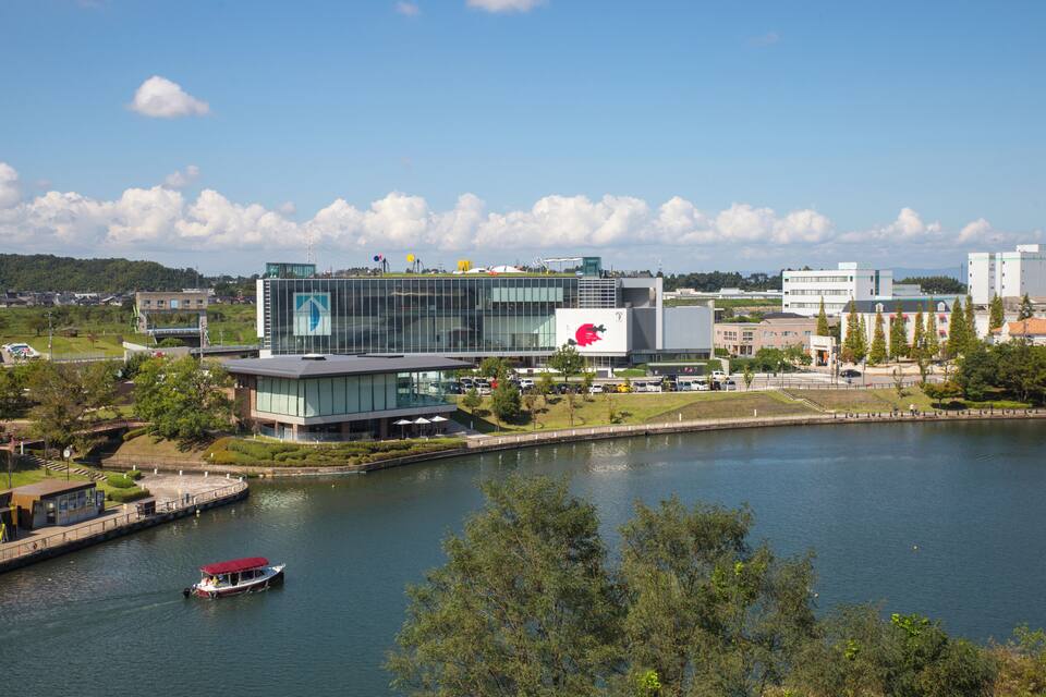Um edifício moderno com fachada de vidro e um logotipo vermelho, localizado próximo a um lago tranquilo com um barco ao primeiro plano.