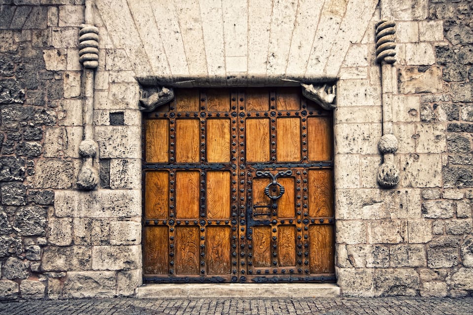 Uma porta de madeira grande com reforços de metal e um batedor, embutida numa parede de pedra.