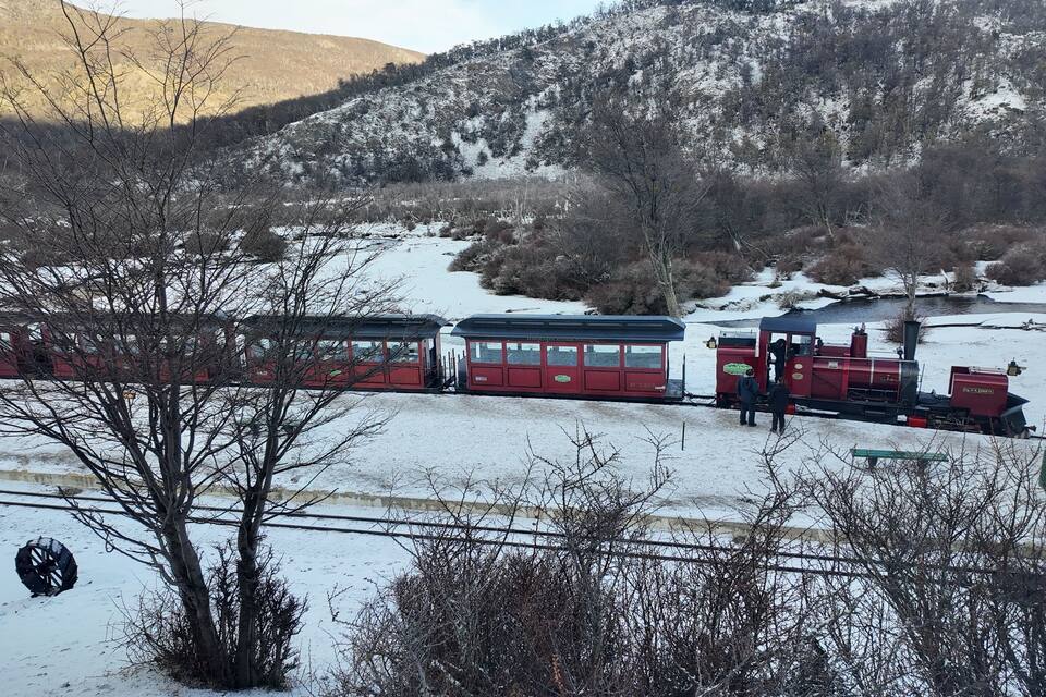 Um trem vermelho sobre trilhos, com o chão coberto de neve e colinas ao fundo.