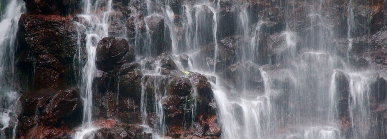 Uma cascata desce sobre rochas escuras, com uma pessoa sentada em uma rocha ao primeiro plano.