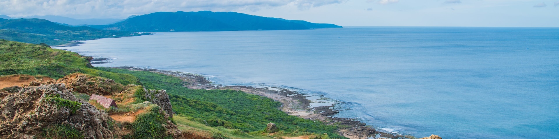 Paisagem costeira com terreno rochoso, área gramada e uma cadeia de montanhas distante sob um céu parcialmente nublado.