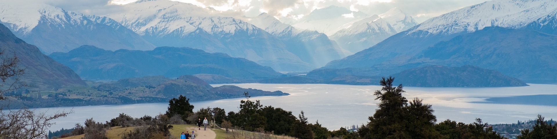 Paisagem montanhosa com um lago e um caminho com pessoas caminhando.