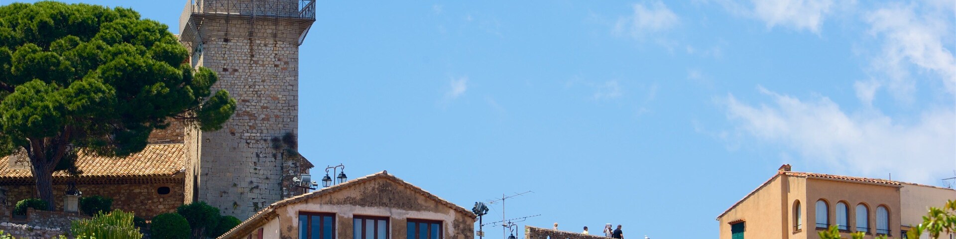 Uma torre do relógio histórica com números romanos, rodeada por arquitetura mediterrânea tradicional e um céu azul límpido.