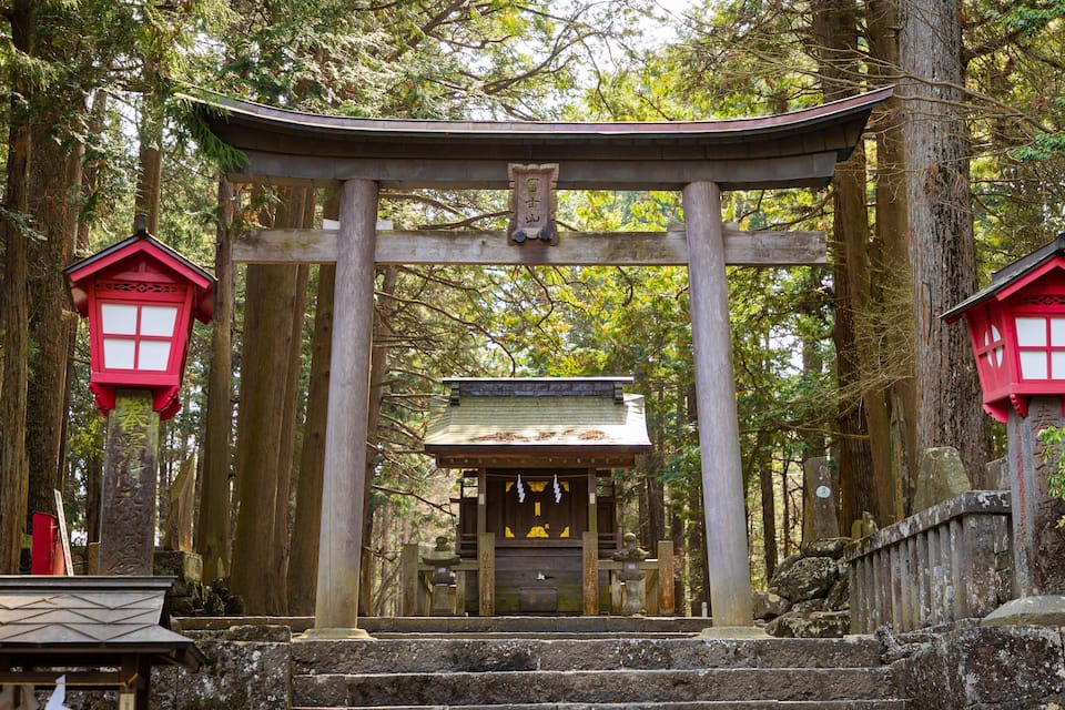 Um tradicional portão torii japonês numa área arborizada, com um pequeno santuário e lanternas.