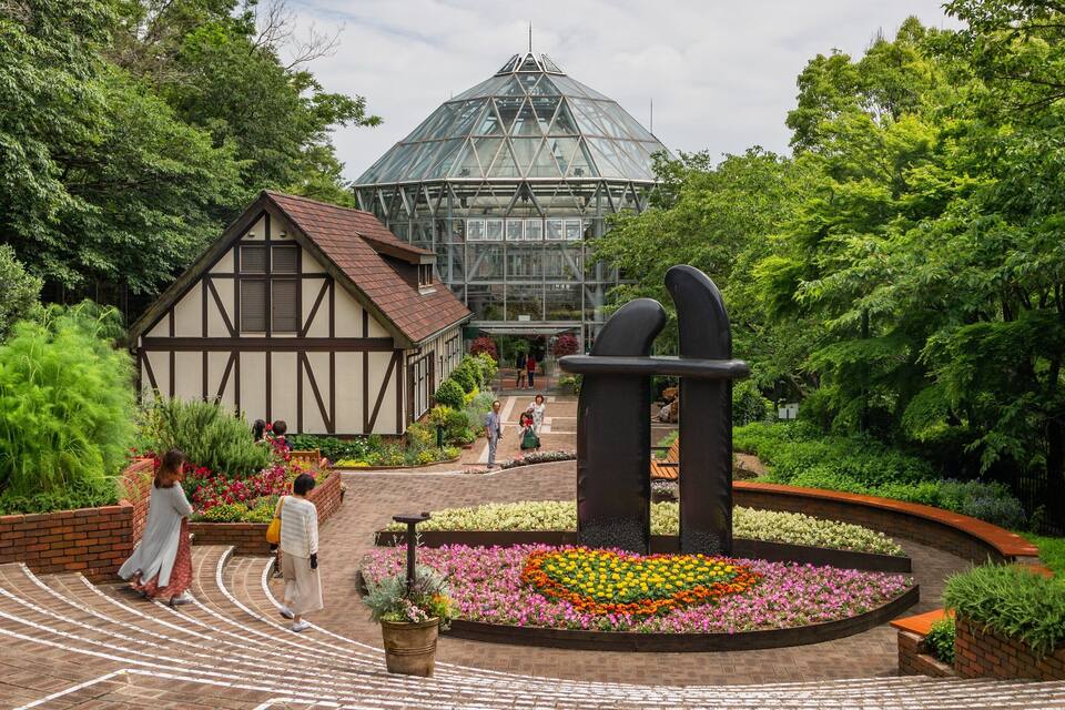 Um jardim com um edifício em estilo Tudor, uma estufa de vidro e uma escultura central.