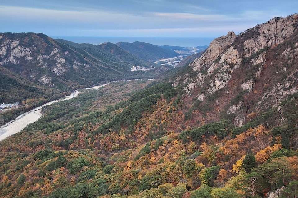 Paisagem montanhosa com folhagem outonal, um rio e edifícios ao longe.