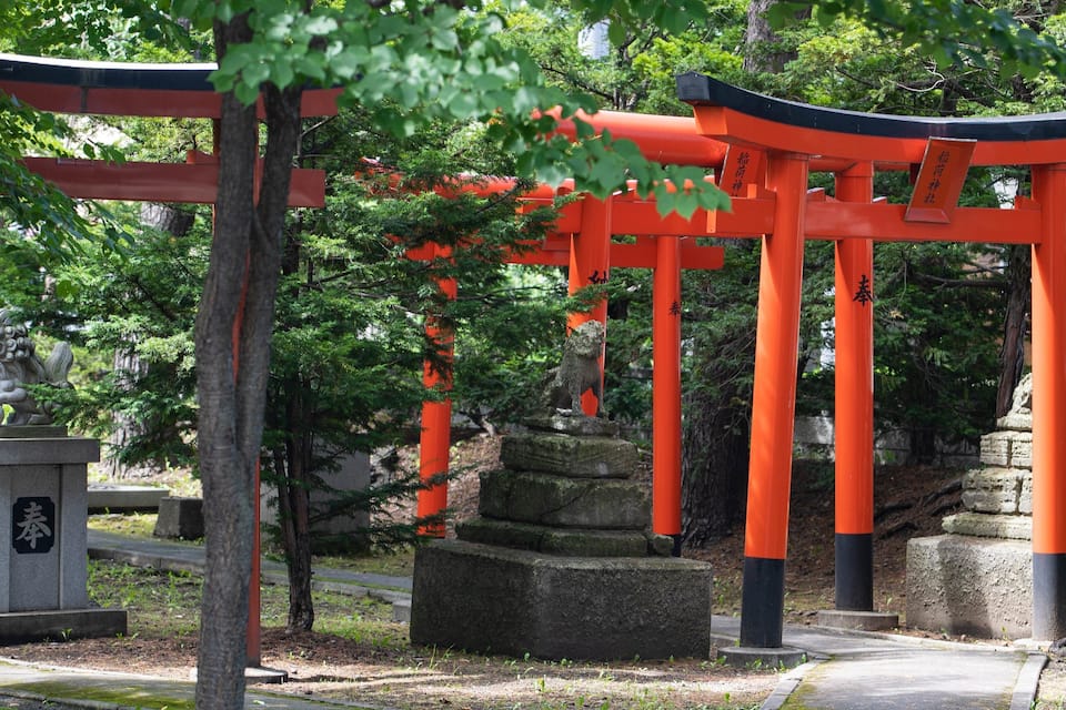 Um tradicional portão torii japonês, com pilares vermelhos e uma estátua de leão de pedra numa área arborizada.