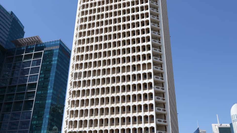 Um edifício alto e branco com fachada em grade, cercado por palmeiras e outros edifícios modernos.