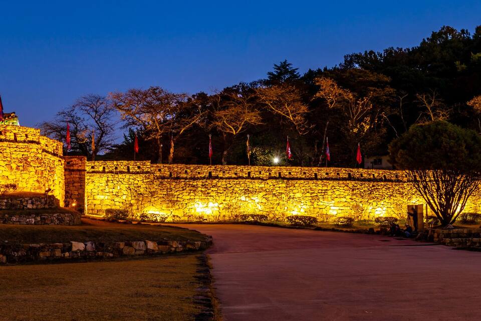 Uma fortaleza de pedra com uma torre redonda, cercada por árvores e iluminada por luzes quentes.