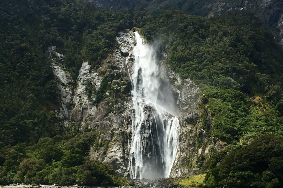 Uma cascata desce por um penhasco rochoso, rodeada por vegetação exuberante e um corpo de água na base.