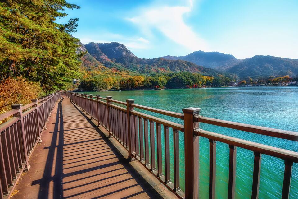 Uma ponte de madeira sobre um lago azul-turquesa, com montanhas ao fundo.