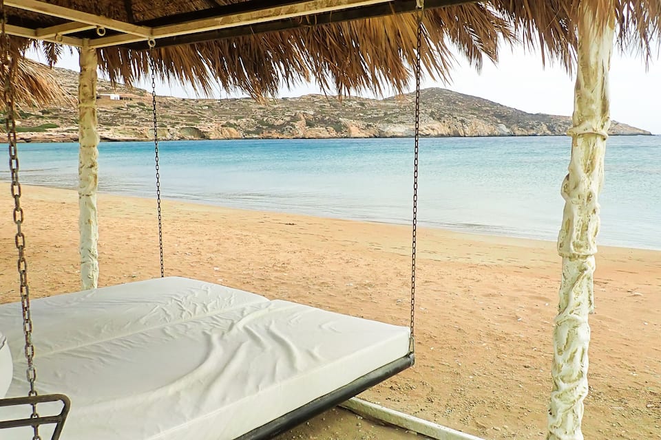 Uma praia com uma rede e um toldo de palha, margem arenosa e montanhas ao fundo.