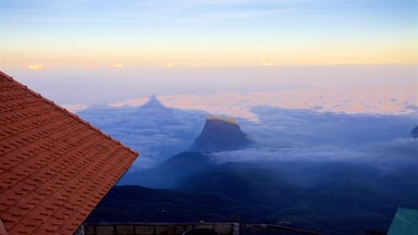 Um pico montanhoso acima das nuvens, uma construção com telhado vermelho e um vale.