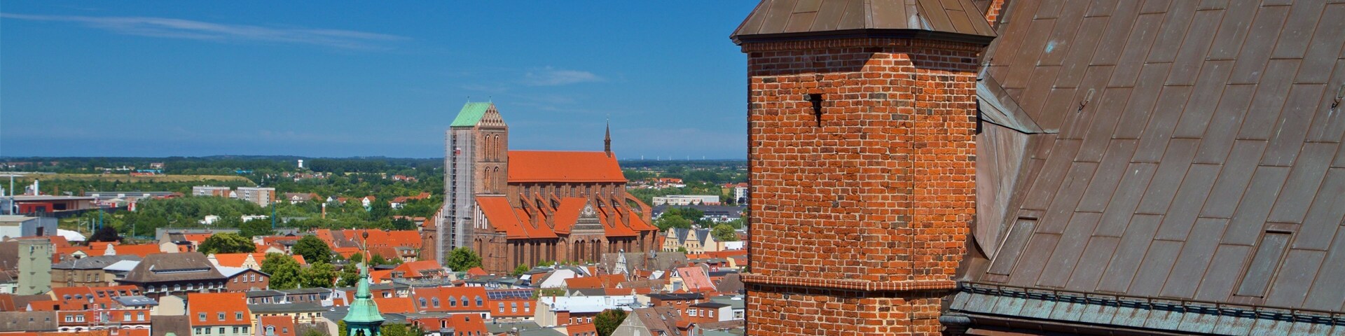 Paisagem urbana com edifícios de telhados vermelhos, uma igreja imponente com uma torre alta e uma torre de tijolos com janelas circulares.