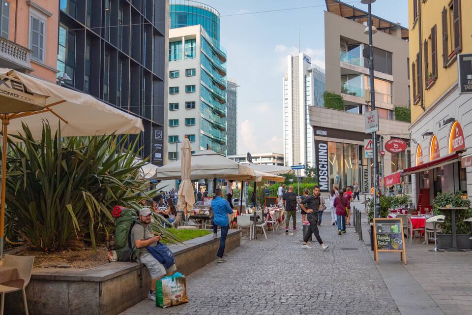 Rua de calçada com cafés ao ar livre, um prédio moderno alto e um edifício histórico com placa indicando pizzaria.