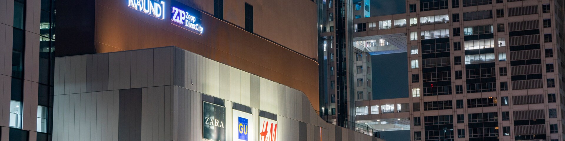 Um edifício moderno com letreiros iluminados para Zara, GUCCI, H&M, ABC-MART e Skechers.