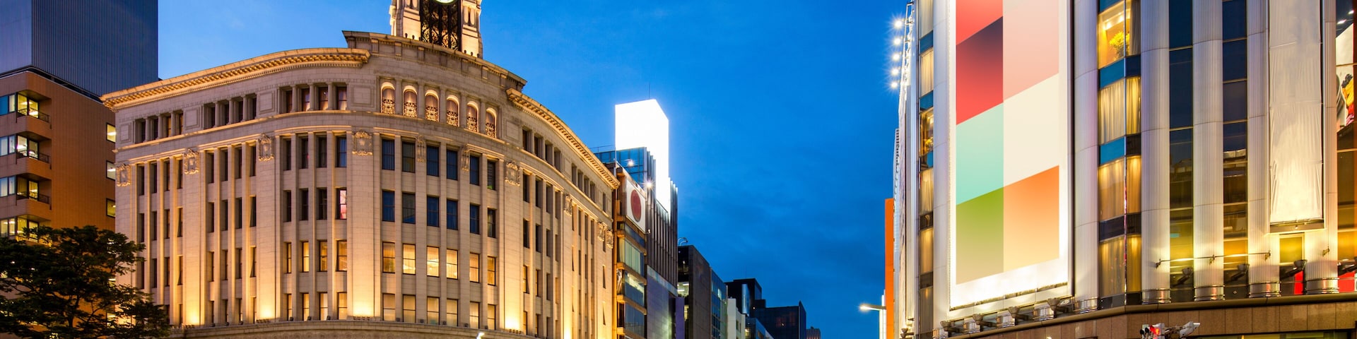 Uma rua urbana ao anoitecer, com edifícios iluminados, uma torre do relógio e um grande outdoor.