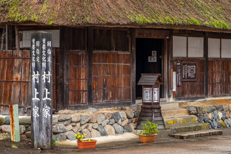 Um edifício tradicional de madeira com telhado de colmo, parede de pedra e uma placa com caracteres japoneses.