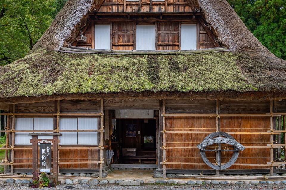 Uma casa tradicional japonesa com telhado de palha, fachada de madeira e um caminho de pedra que leva à entrada.