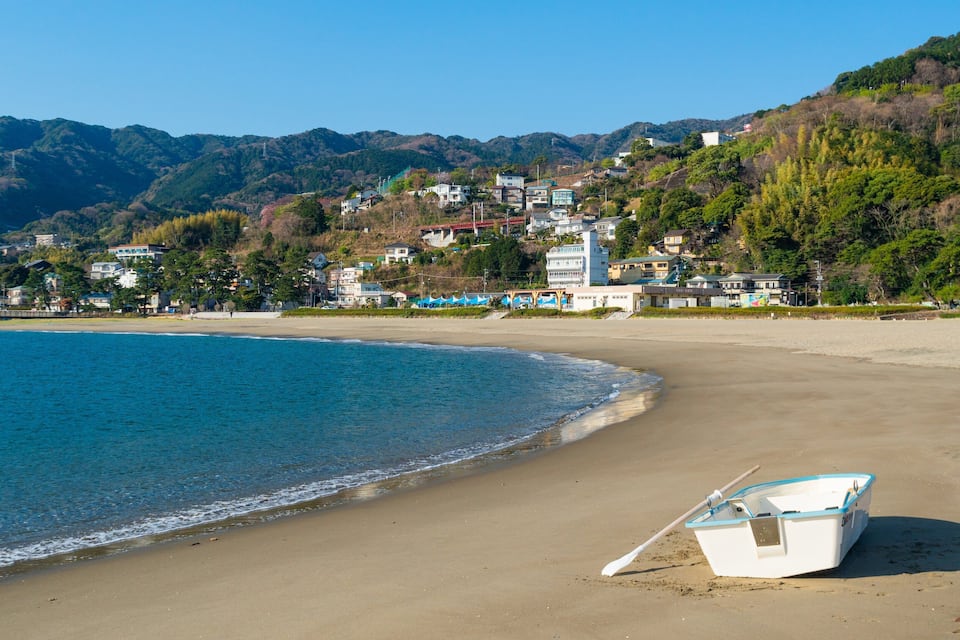 Uma praia com um barco na areia, mar azul cristalino e casas numa encosta ao fundo.