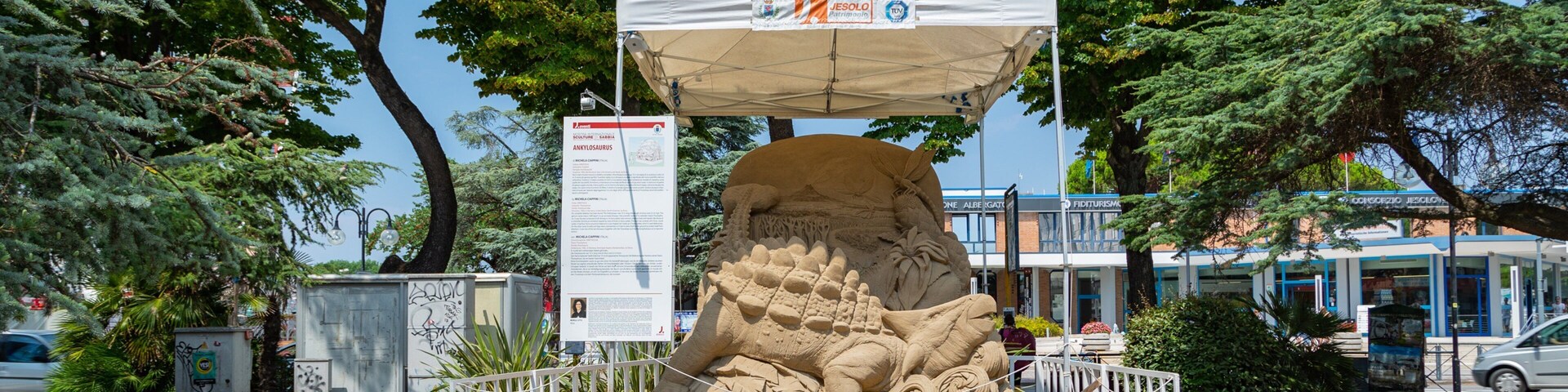 Grande escultura de areia de dinossauro em parque, com uma tenda e placas informativas.