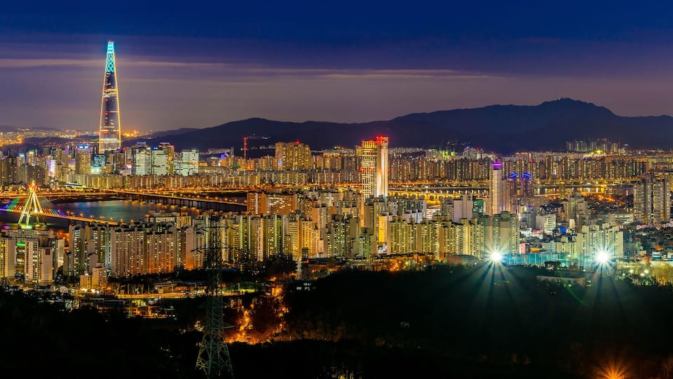 Vista urbana noturna com edifícios iluminados, uma torre imponente e um rio.