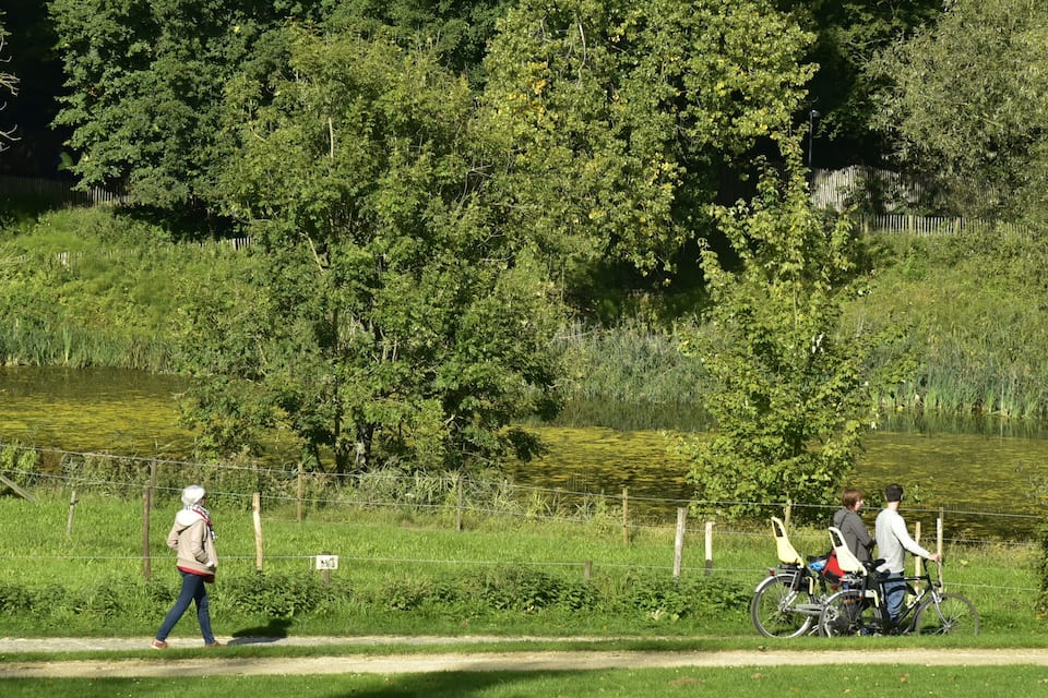 Uma pessoa caminhando por um caminho ao lado de um lago, com uma bicicleta estacionada perto e uma cerca ao fundo.