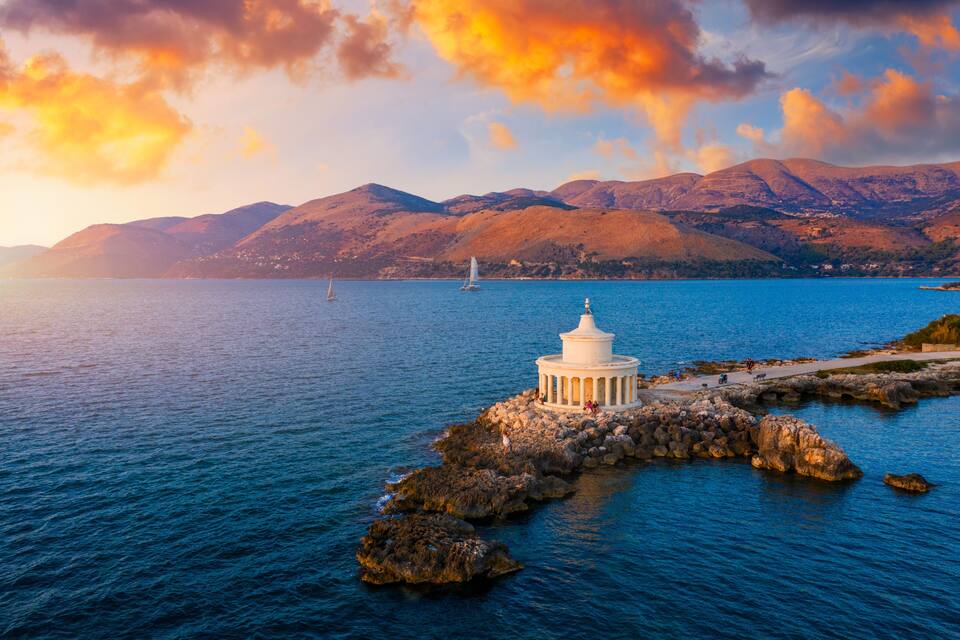 Uma paisagem costeira com um farol branco em um afloramento rochoso, mar azul calmo e montanhas ao fundo.