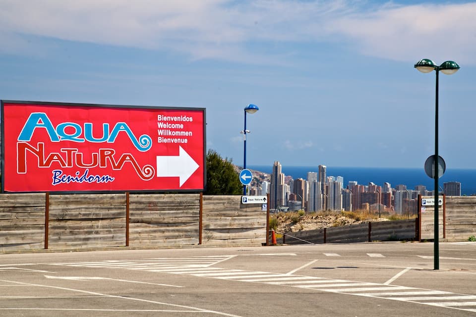 Uma placa vermelha com 'AQUA NATURA Benidorm' e uma seta apontando para a direita. Ao fundo, vista urbana com edifícios e o mar.