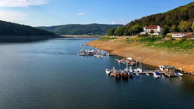 Vista para o lago com barcos atracados num cais, um restaurante e uma colina com edifícios ao fundo.