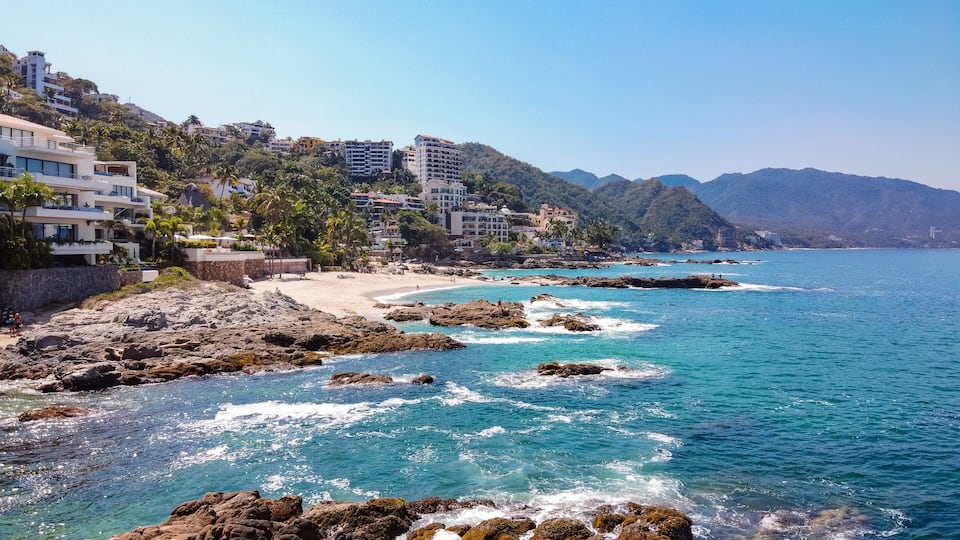 Paisagem costeira com costas rochosas, água azul cristalina e edifícios numa encosta.