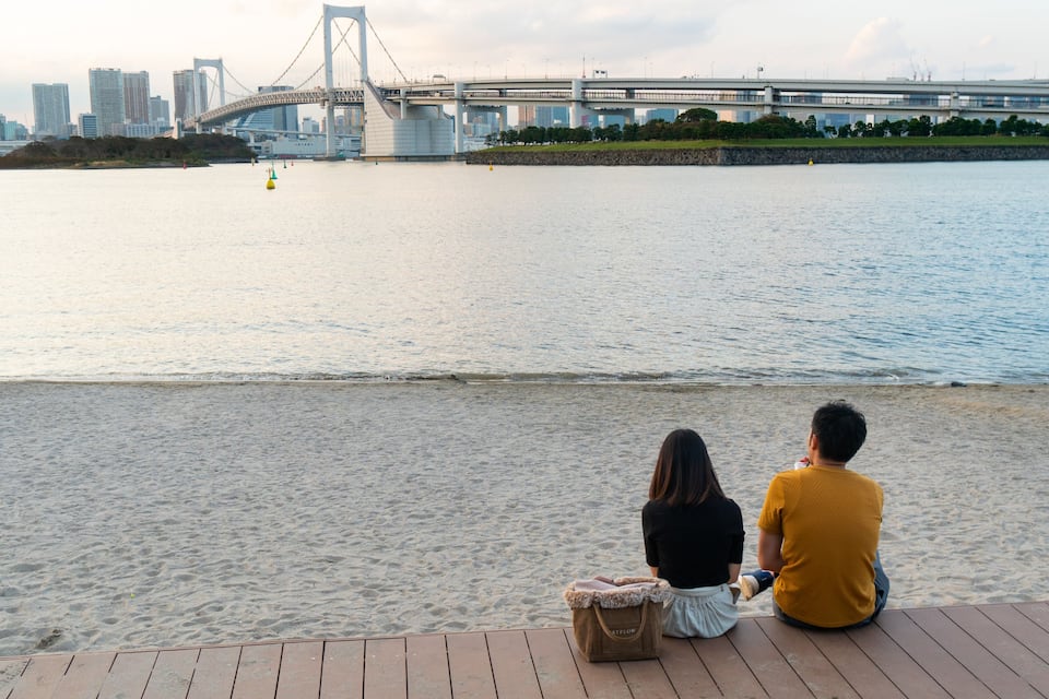 Um casal sentado em um deck de madeira à beira de um rio, com vista para uma ponte e o horizonte da cidade.