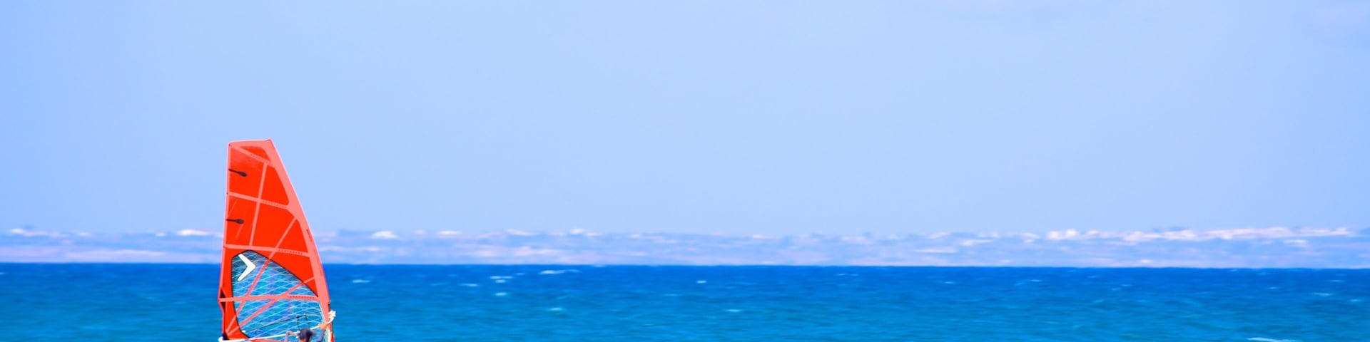 Uma pessoa praticando windsurf em um mar azul e límpido com uma vela vermelha.
