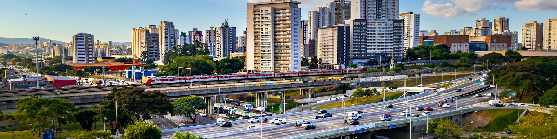 Paisagem urbana com um trem em destaque em uma linha elevada, vários edifícios altos e uma rodovia movimentada.