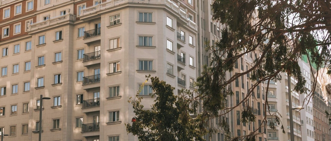Um edifício de vários andares com cúpula e varandas, uma rua movimentada com carros e pedestres, e árvores em primeiro plano.