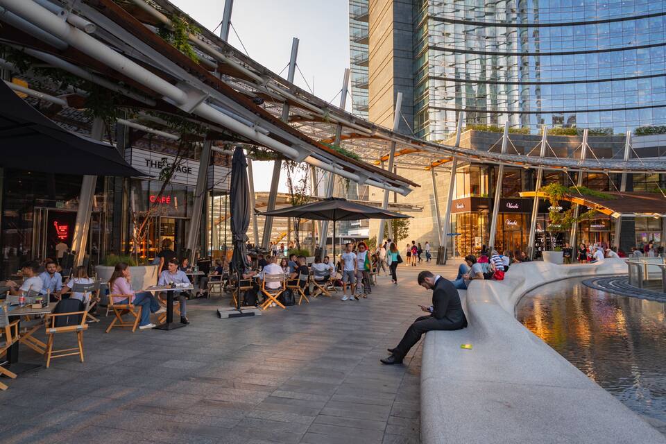 Uma área de refeições ao ar livre com mesas e cadeiras, uma fonte e edifícios modernos ao fundo.