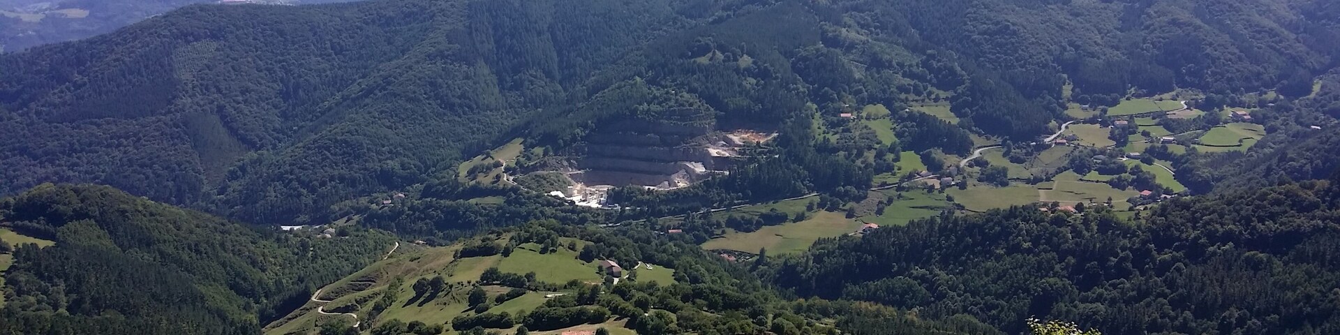 Um vale com um lago rodeado por colinas florestadas e montanhas ao longe.