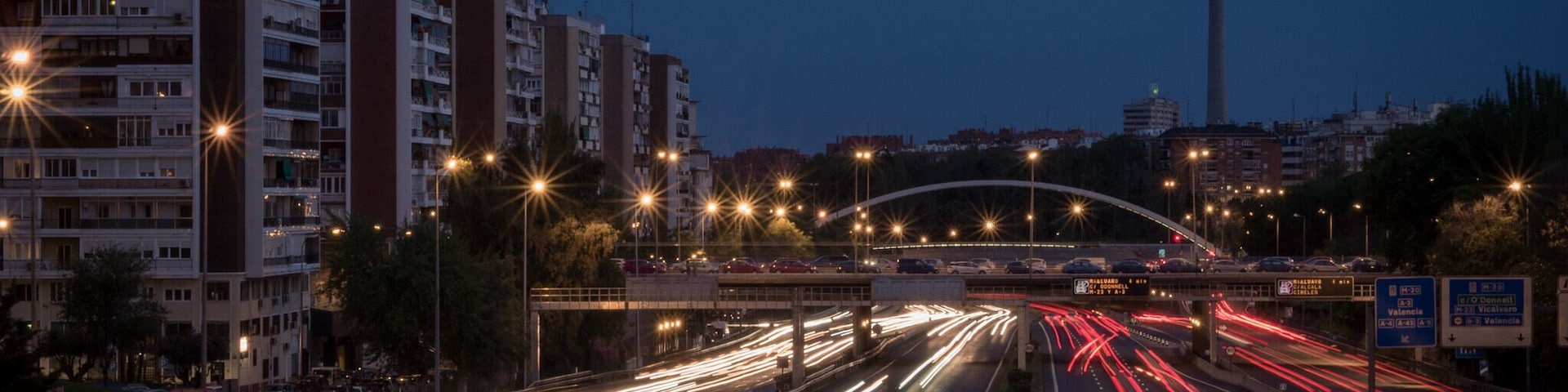 Autoestrada com várias faixas, rastros de luz, uma ponte e uma torre ao fundo.