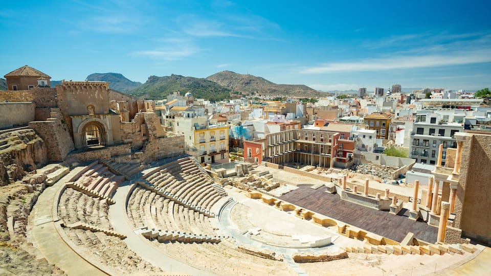 Um antigo anfiteatro de pedra com degraus e palco central, rodeado por edifícios modernos e uma paisagem montanhosa.