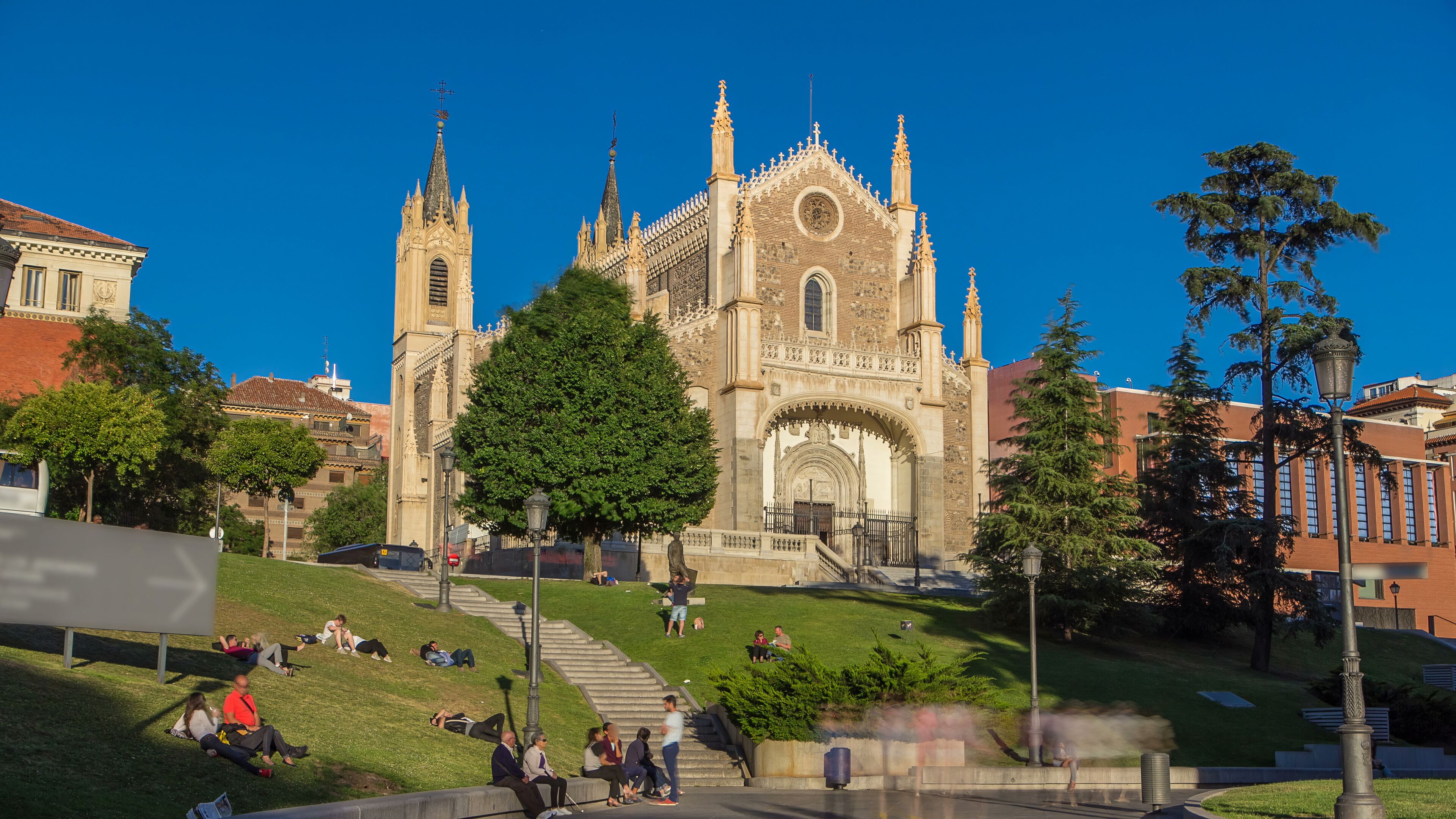 Uma catedral histórica com arquitetura detalhada, uma árvore grande em primeiro plano e pessoas relaxando na grama.