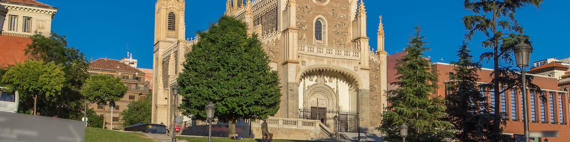Uma catedral histórica com arquitetura detalhada, uma árvore grande em primeiro plano e pessoas relaxando na grama.