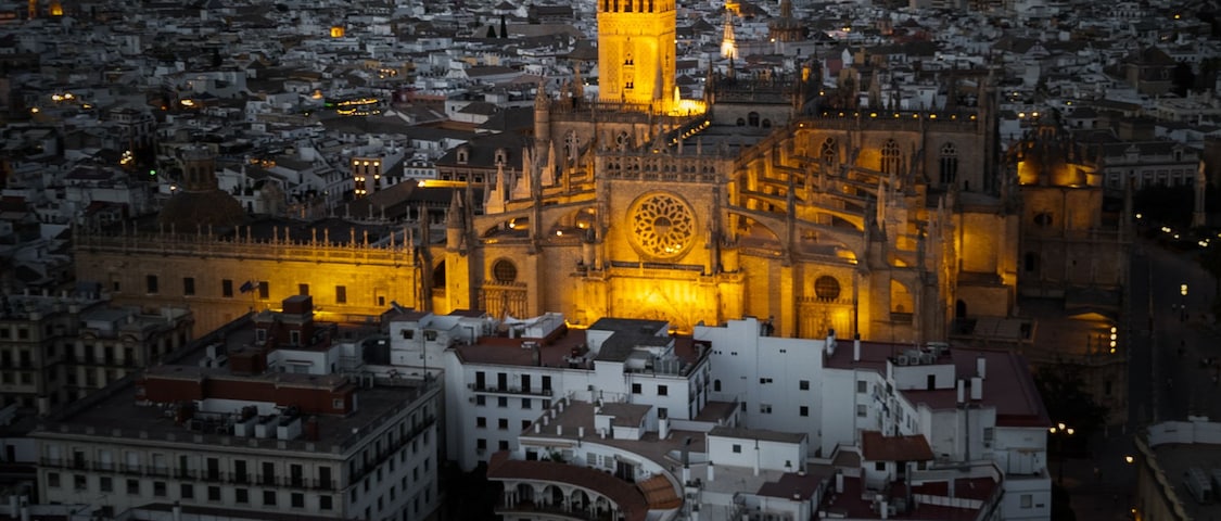 Paisagem urbana ao entardecer, com uma catedral iluminada e uma torre alta ao fundo.
