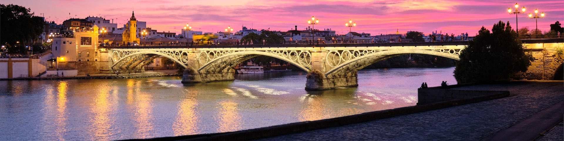 Uma ponte de pedra sobre um rio, com edifícios da cidade ao fundo e um céu rosado ao entardecer.