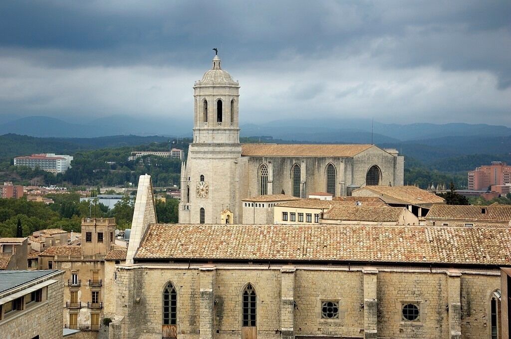 Uma igreja histórica com uma torre de sino e um relógio, rodeada por edifícios urbanos e montanhas ao fundo.
