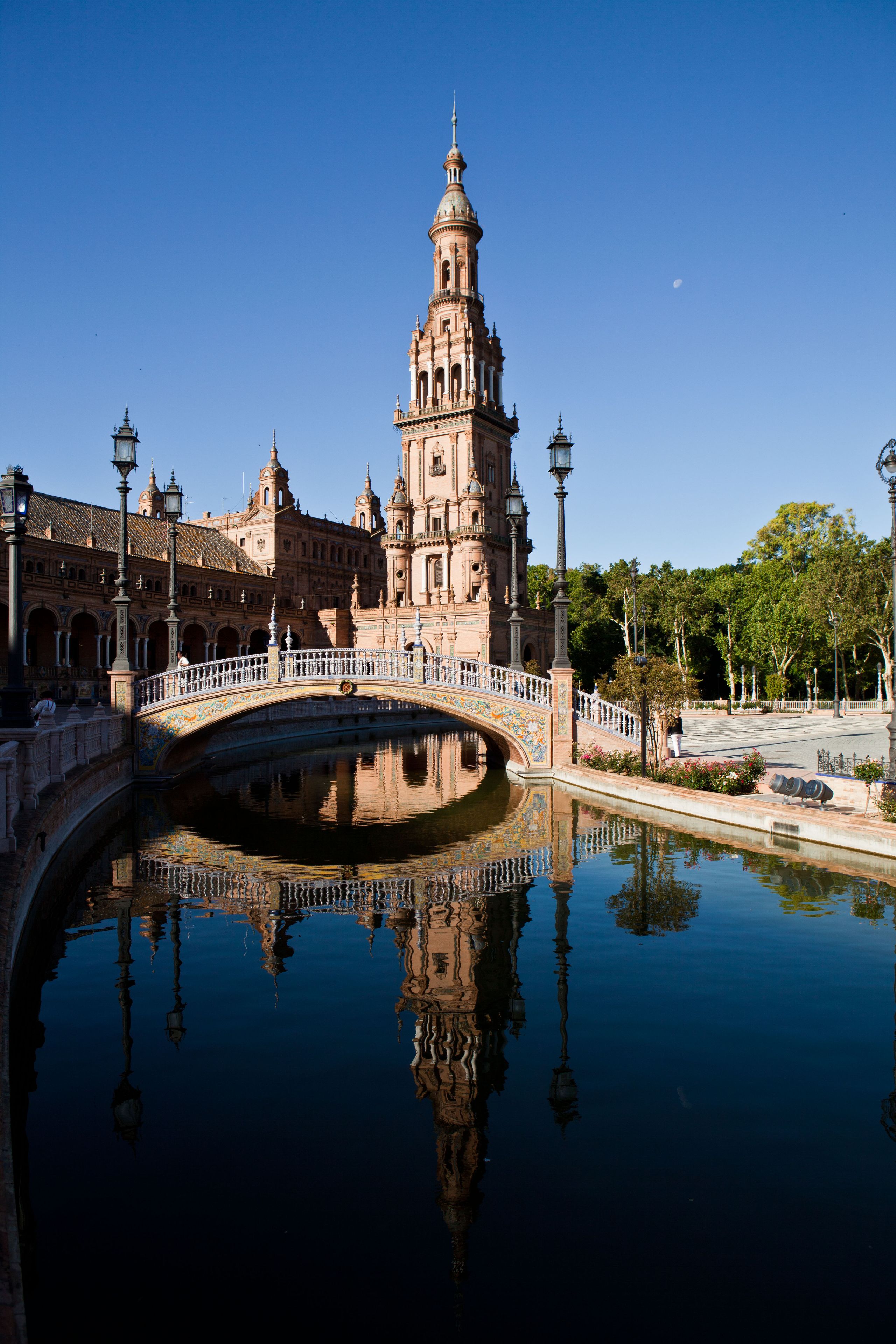 Um edifício histórico com uma torre imponente, uma ponte decorativa e um corpo d'água calmo que reflete a arquitetura.
