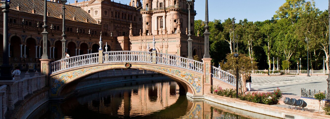 Um edifício histórico com uma torre imponente, uma ponte decorativa e um corpo d'água calmo que reflete a arquitetura.